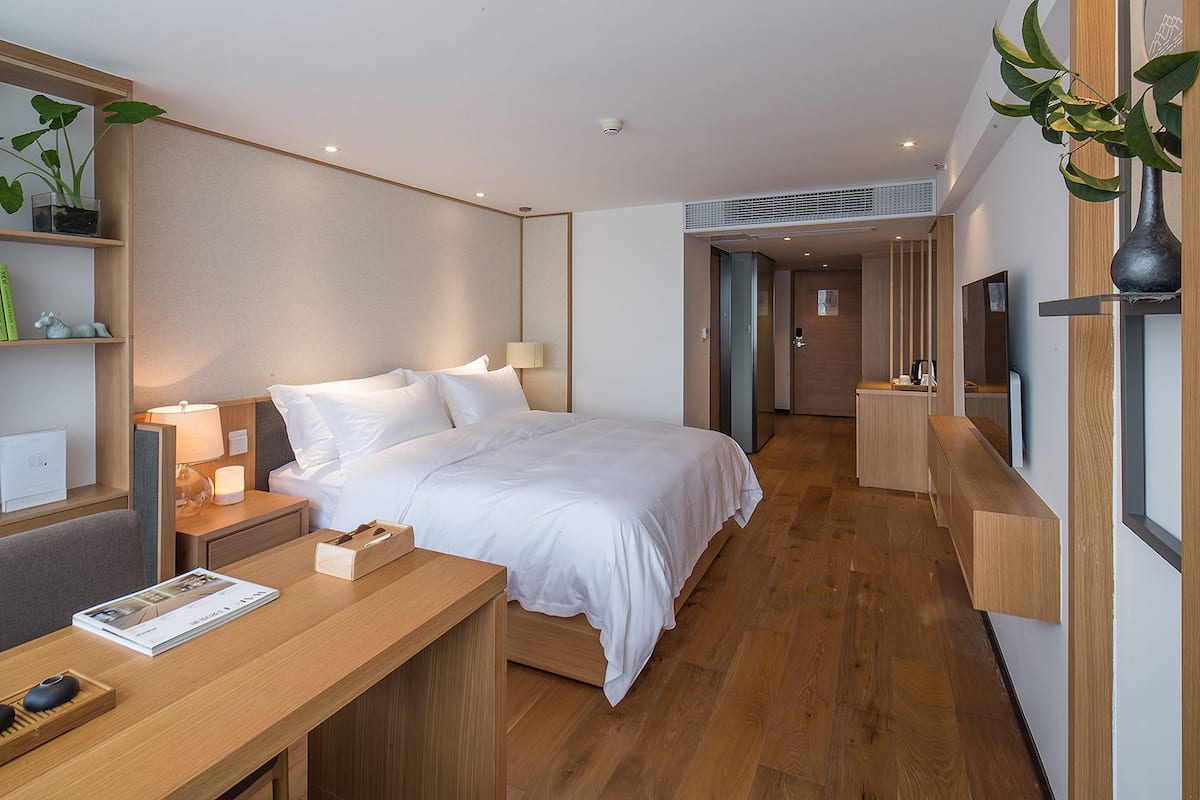 Quarto de hotel moderno com uma escrivaninha de madeira, uma cama com roupa branca, uma mesa de cabeceira com abajur e uma TV em um suporte de madeira.