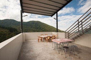 Varanda coberta ao ar livre com mesa e cadeiras, escada e vista para uma montanha arborizada.
