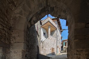 Um edifício de pedra com telhado inclinado e uma pequena varanda, visto através de uma porta de pedra em arco.