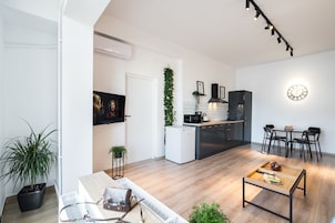 Cozinha moderna com armários brancos, bancada preta e forno embutido. Possui uma área de refeições com mesa e cadeiras de madeira, e uma sala de estar com sofá e mesa de centro.