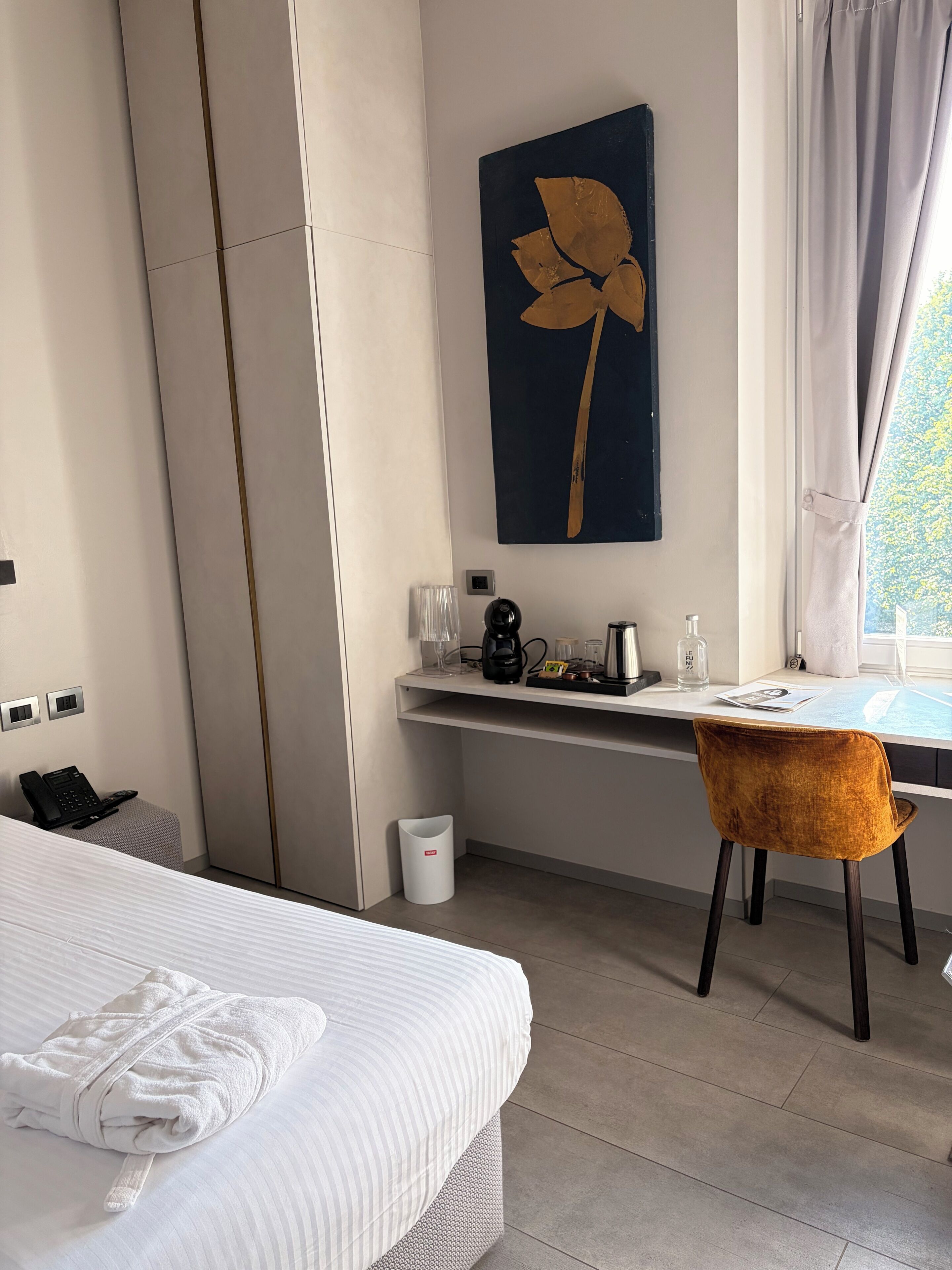 Quarto de hotel com uma cama, uma escrivaninha com cadeira, uma cafeteira e um quadro na parede.