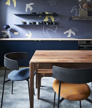 Cozinha moderna com paredes azul-escuras, armários de madeira e forno embutido. Há duas cadeiras com assentos azul e laranja, e uma mesa de madeira.