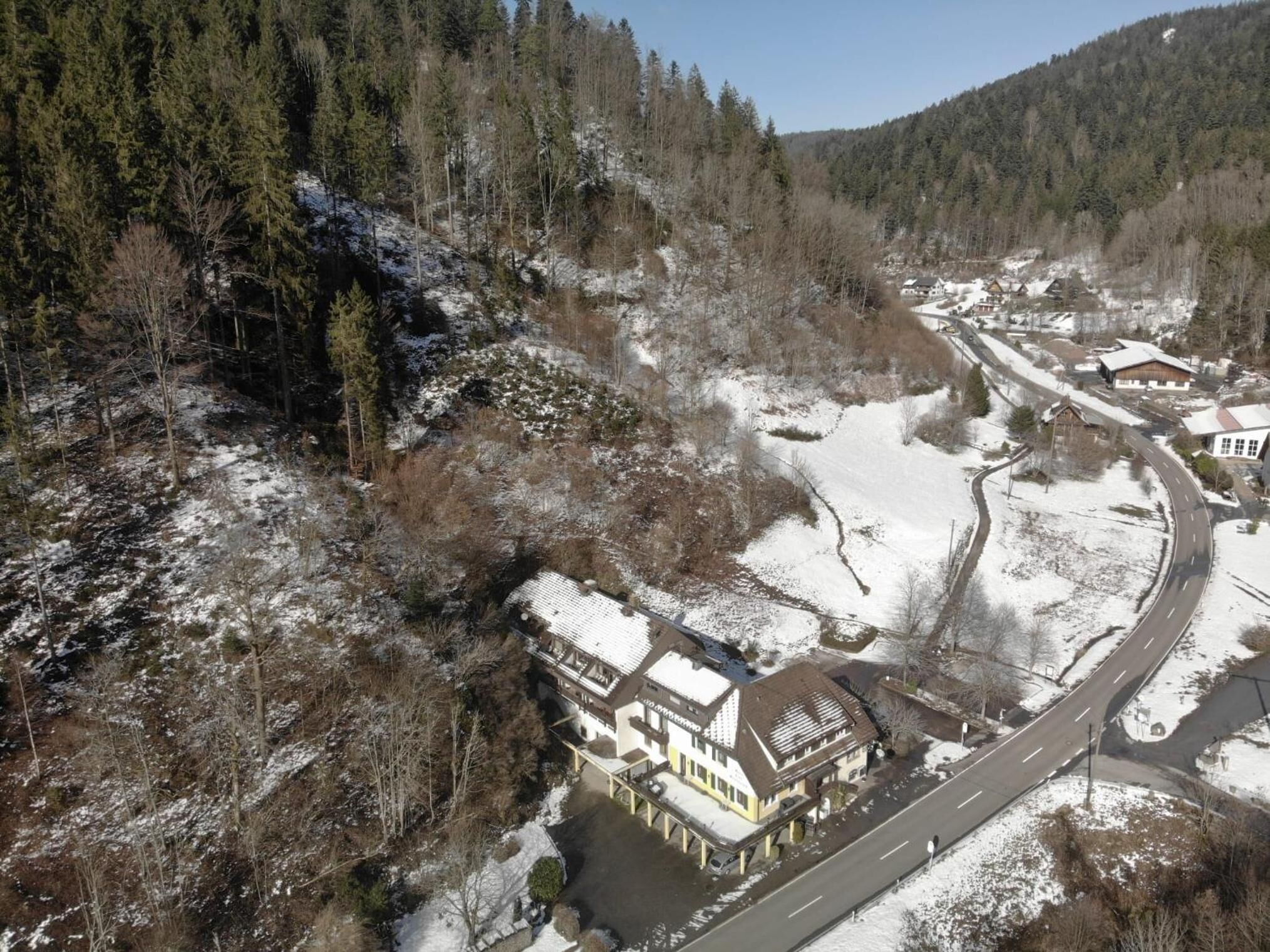 Paisagem coberta de neve com uma estrada serpenteando por uma área arborizada, levando a um edifício com telhado inclinado e entrada visível.