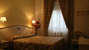 Quarto de hotel com duas camas, mesa de cabeceira, abajur, vaso com flores e uma cadeira.