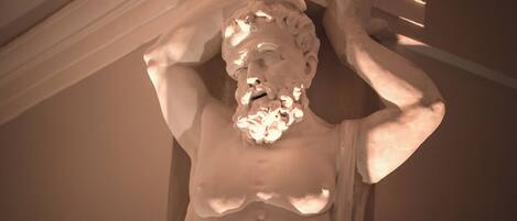 Escultura de uma figura com braços levantados, em um espaço com teto inclinado e vigas aparentes.