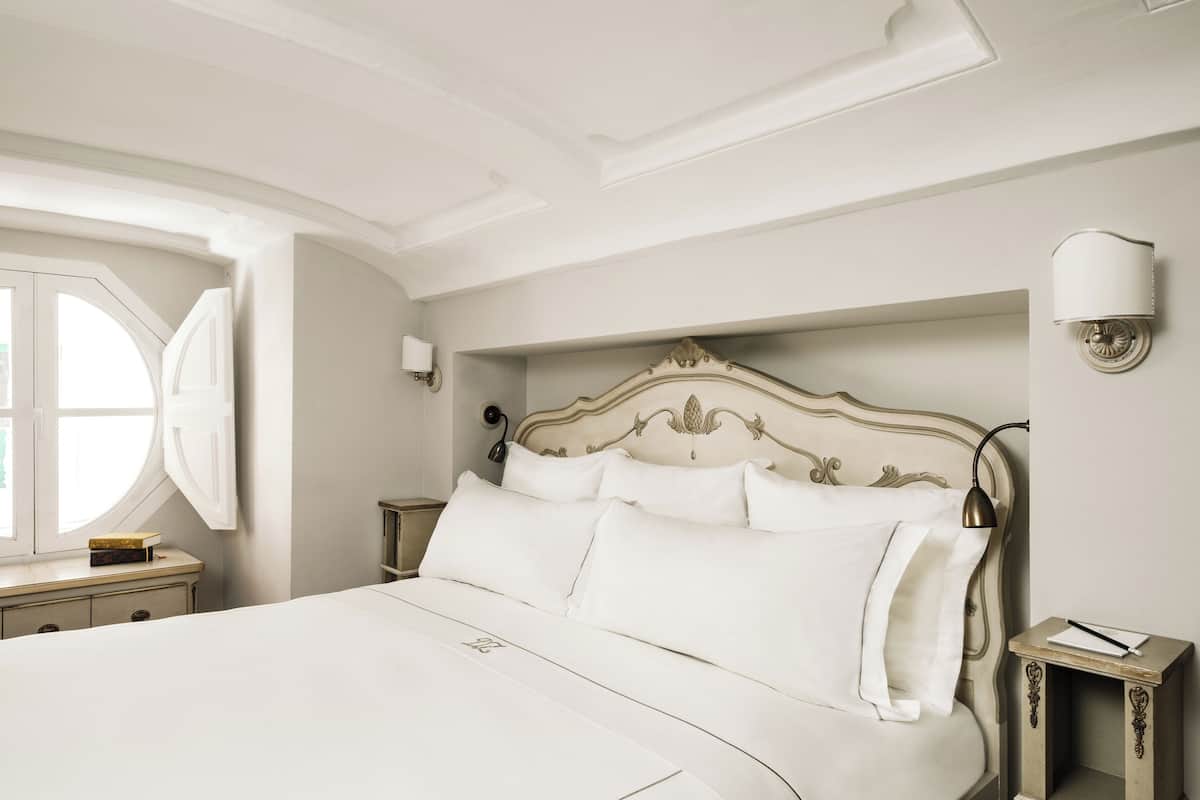 Cama bem arrumada com cabeceira, mesa de cabeceira com abajur e janela com cortinas.