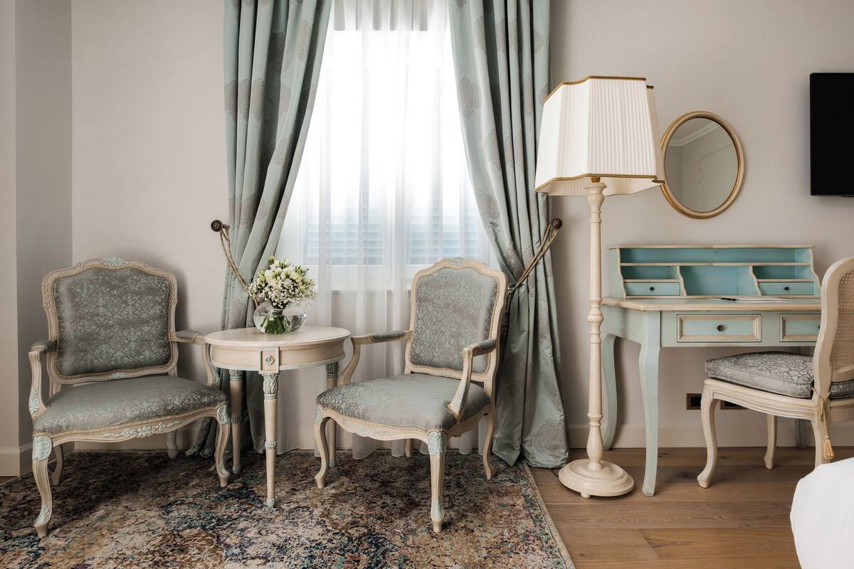 Quarto com duas poltronas, uma mesa redonda pequena com um vaso de flores, uma luminária, um penteadeira e um espelho na parede.