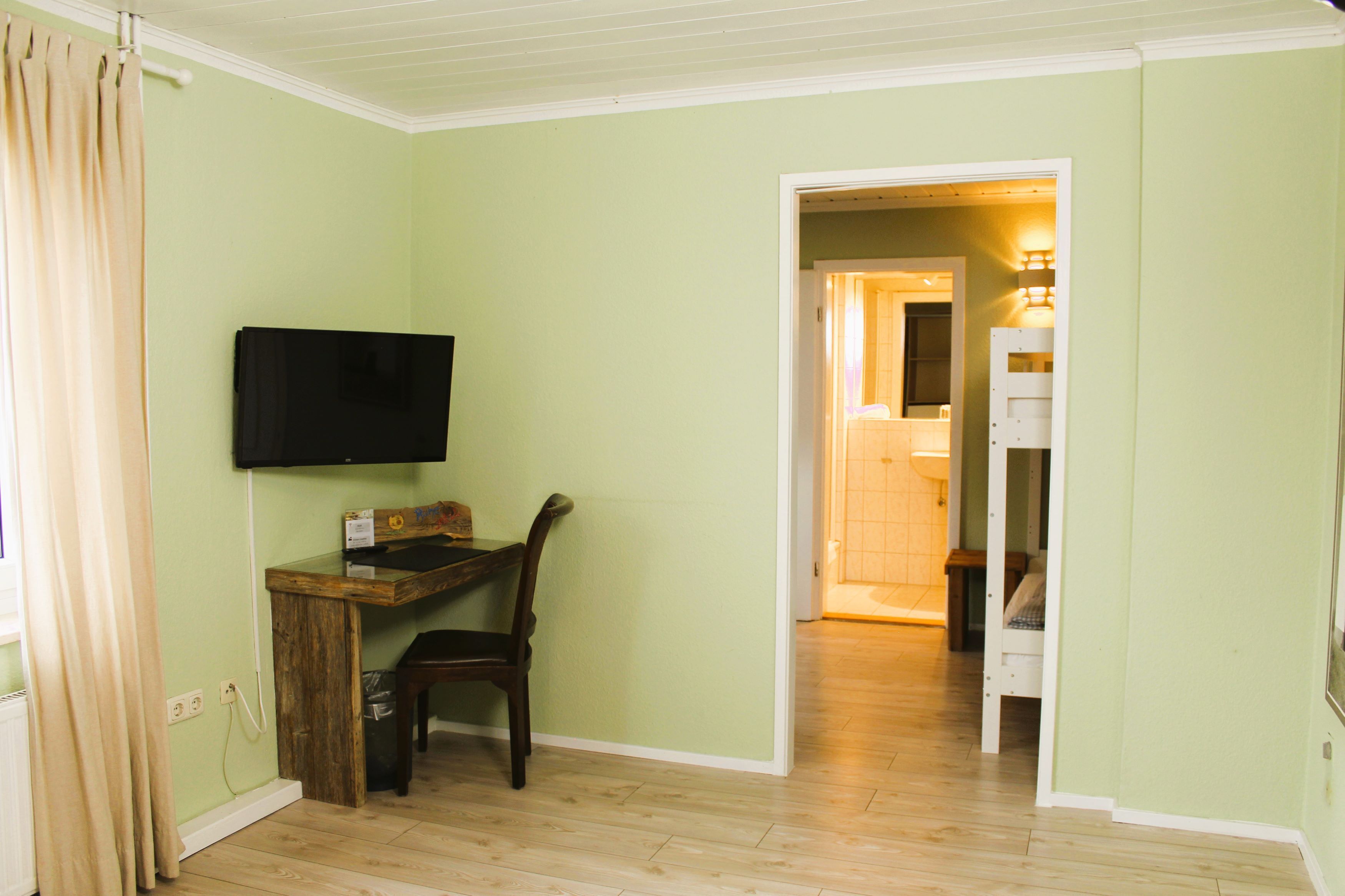 Quarto com escrivaninha de madeira, uma cadeira, televisão fixada na parede e porta que dá para outro aposento.