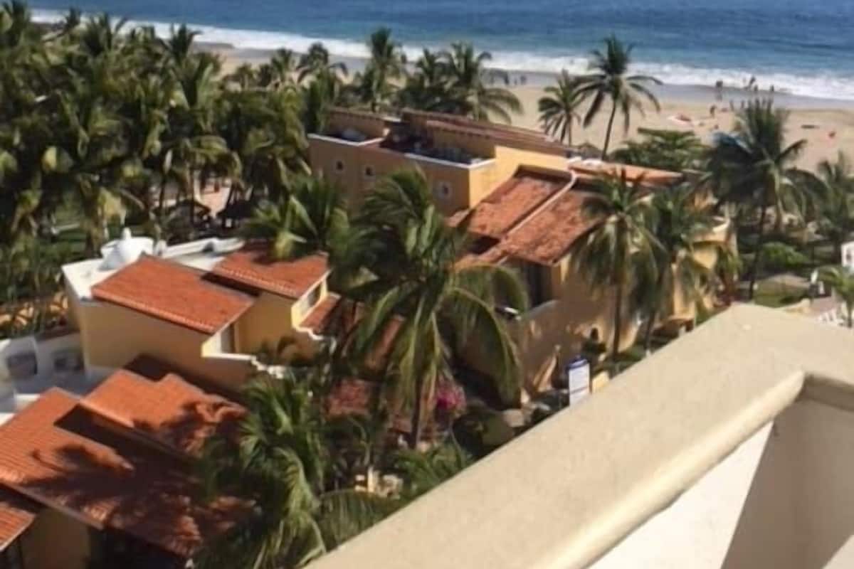 Vista costeira com praia, palmeiras e edifícios com telhados de telha vermelha.