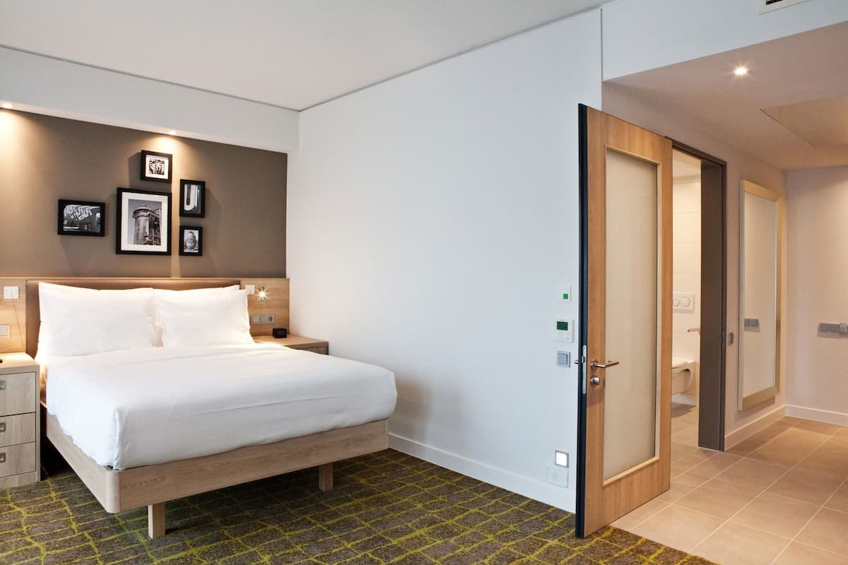 Quarto de hotel com cama, mesas de cabeceira, luminária na parede, porta e tapete estampado.