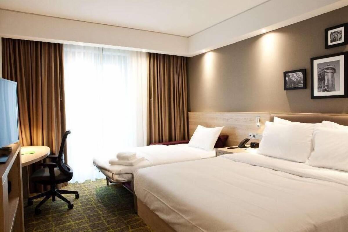 Quarto de hotel com duas camas, uma escrivaninha com cadeira, televisão e janela com cortinas.