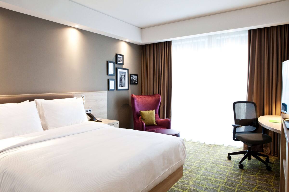 Quarto de hotel com uma cama grande, uma escrivaninha com cadeira, uma poltrona vermelha e uma janela ampla com cortinas.