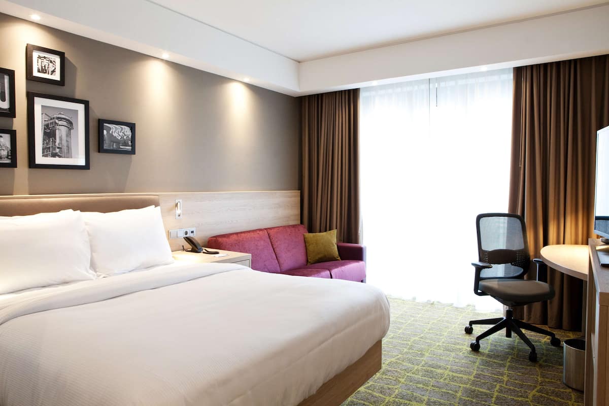 Quarto de hotel com uma cama grande, uma escrivaninha com cadeira, um pequeno sofá e quadros nas paredes.