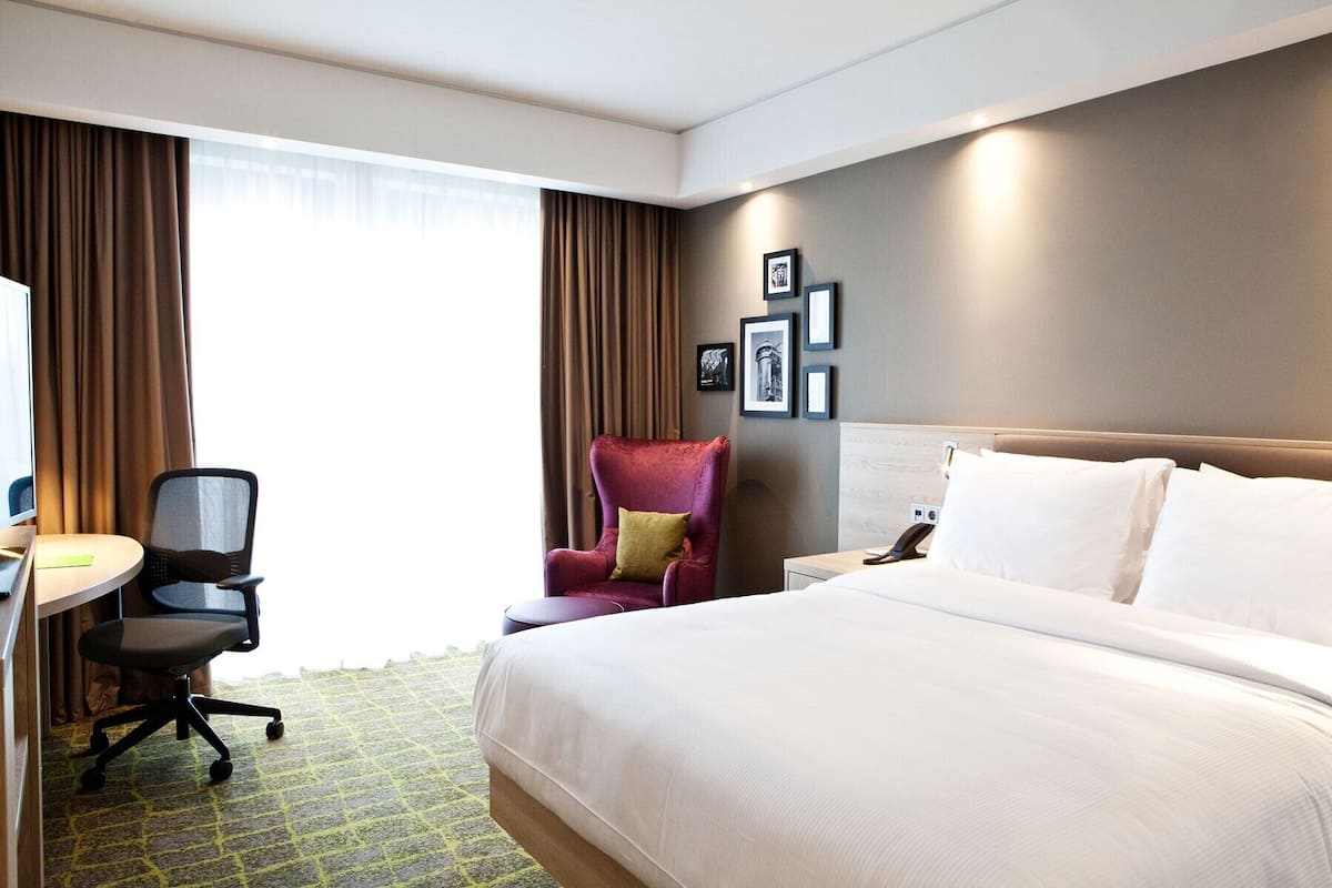 Quarto de hotel com cama, secretária, cadeira e uma janela grande com cortinas.