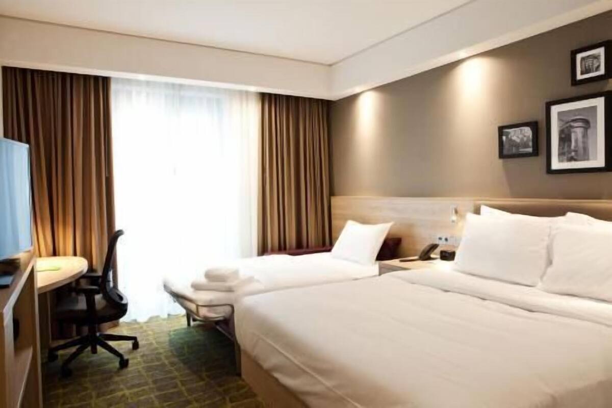 Quarto de hotel com duas camas, uma escrivaninha, uma cadeira, televisão e janela com cortinas.