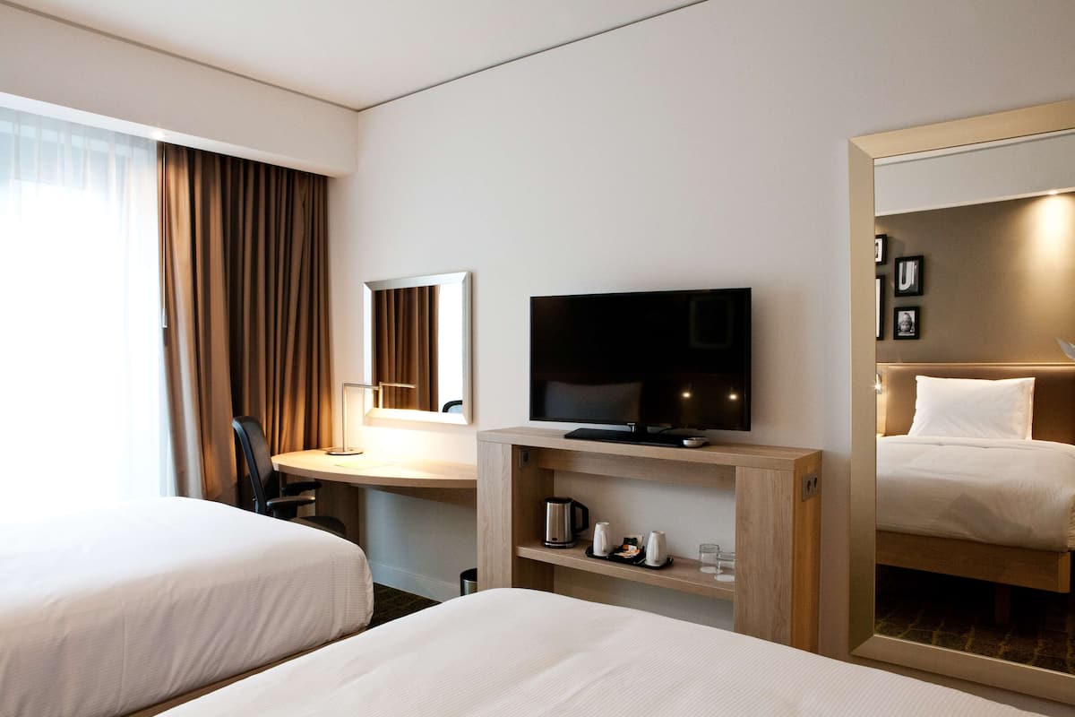 Quarto de hotel com duas camas, uma mesa de trabalho, televisão, máquina de café e espelho.