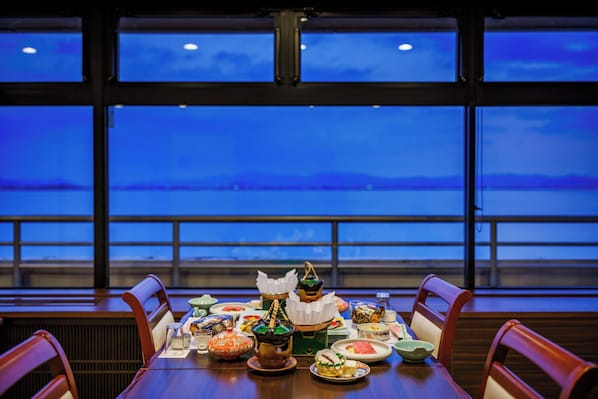 Mesa de jantar posta com diversos pratos e bules, com vista para um corpo de água através de grandes janelas.