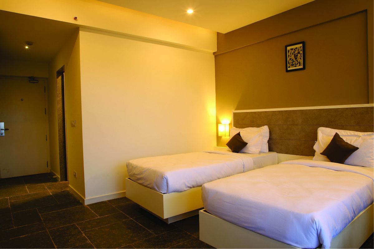 Quarto de hotel com duas camas de solteiro, cabeceira, luminária fixada na parede e porta que dá para outro quarto.