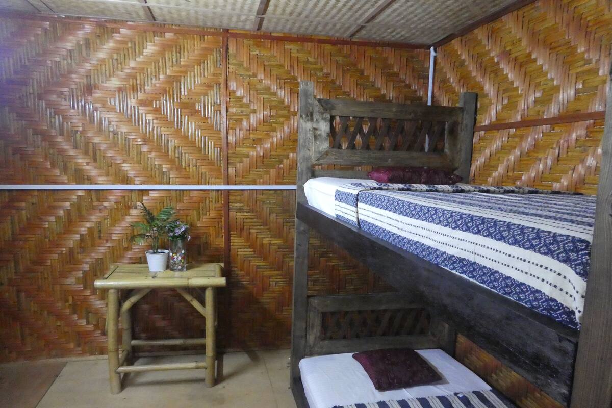 Quarto com uma cama de solteiro, uma pequena mesa de madeira com uma planta e um painel de bambu trançado.