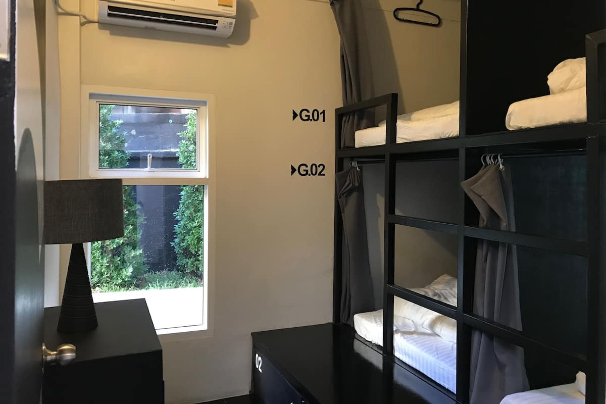 Um quarto em dormitório com beliches, ar condicionado, janela com vista para vegetação e uma escrivaninha preta com luminária.