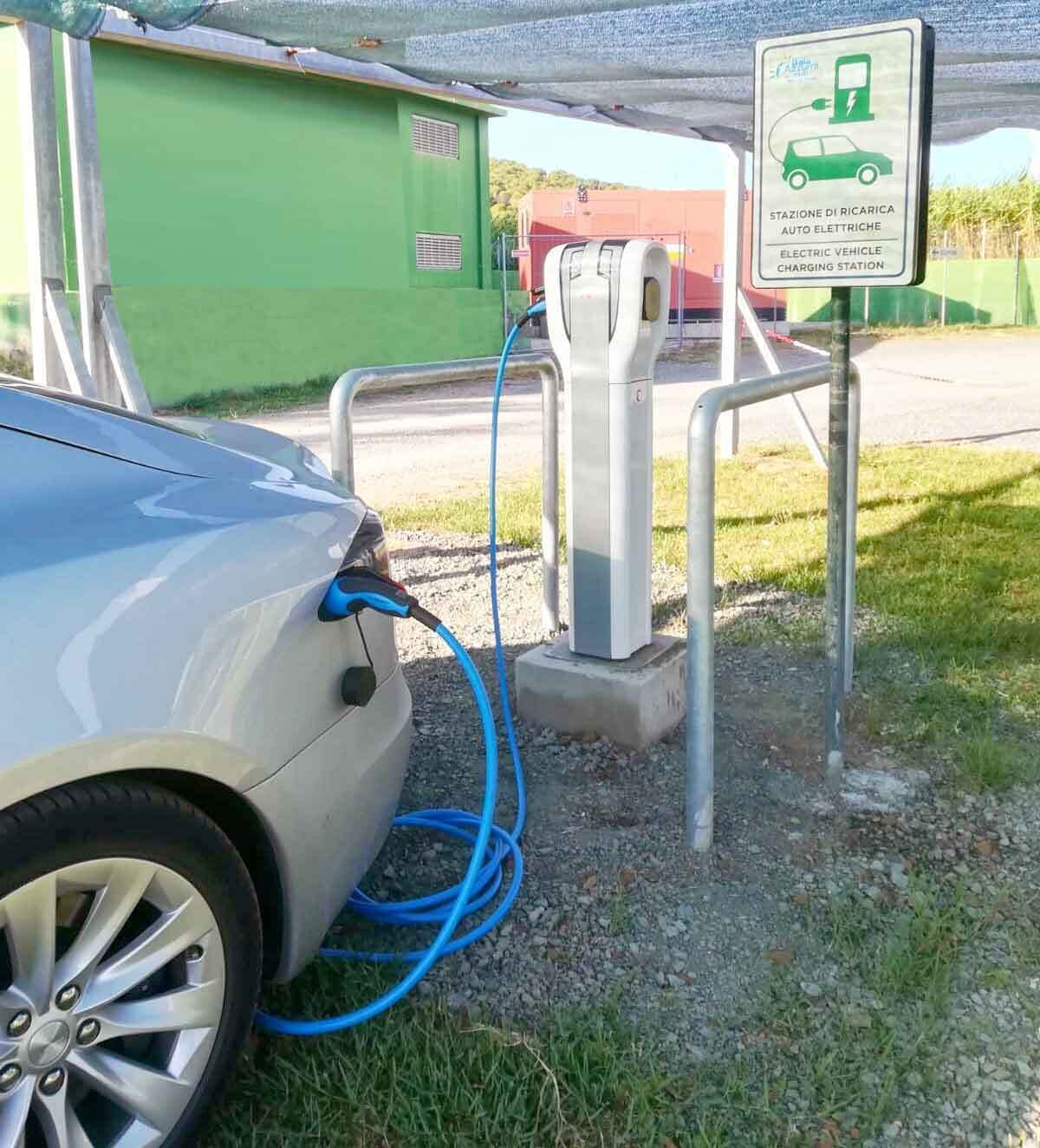 Um carro conectado a uma estação de carregamento para veículos elétricos, com uma placa indicando ser uma estação RICARICA AUTO ELECTRICHE.