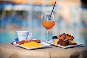Mesa de café da manhã com prato de ovos, bacon e torradas, um copo de suco de laranja e uma xícara de café, à beira de uma piscina.