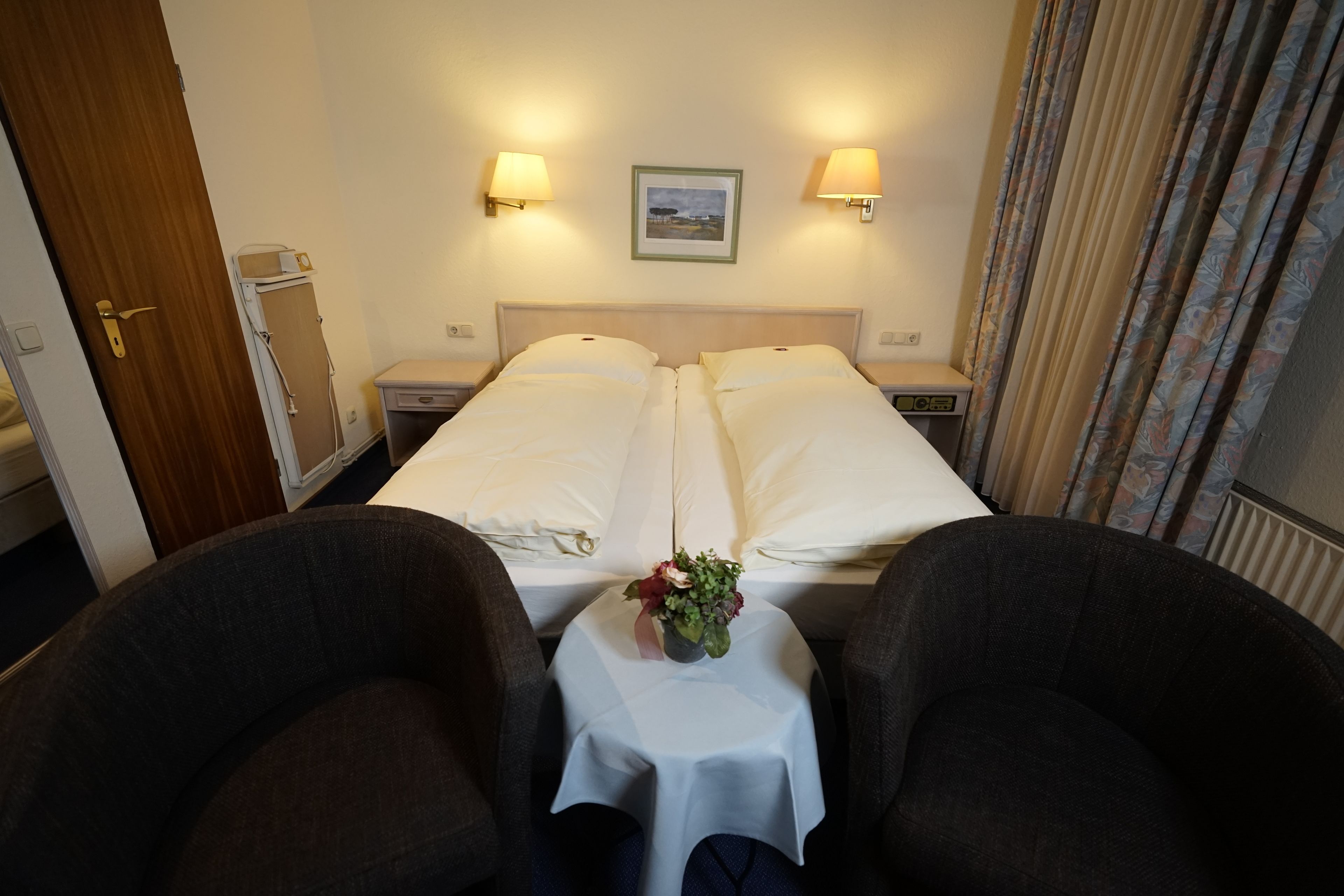 Quarto de hotel com uma cama de casal, duas mesas de cabeceira, duas luminárias, uma mesinha com uma planta e uma porta.