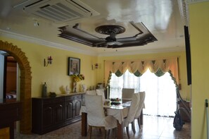 Uma sala de jantar com uma mesa posta para quatro pessoas, um ventilador de teto e vista pela janela com cortinas.