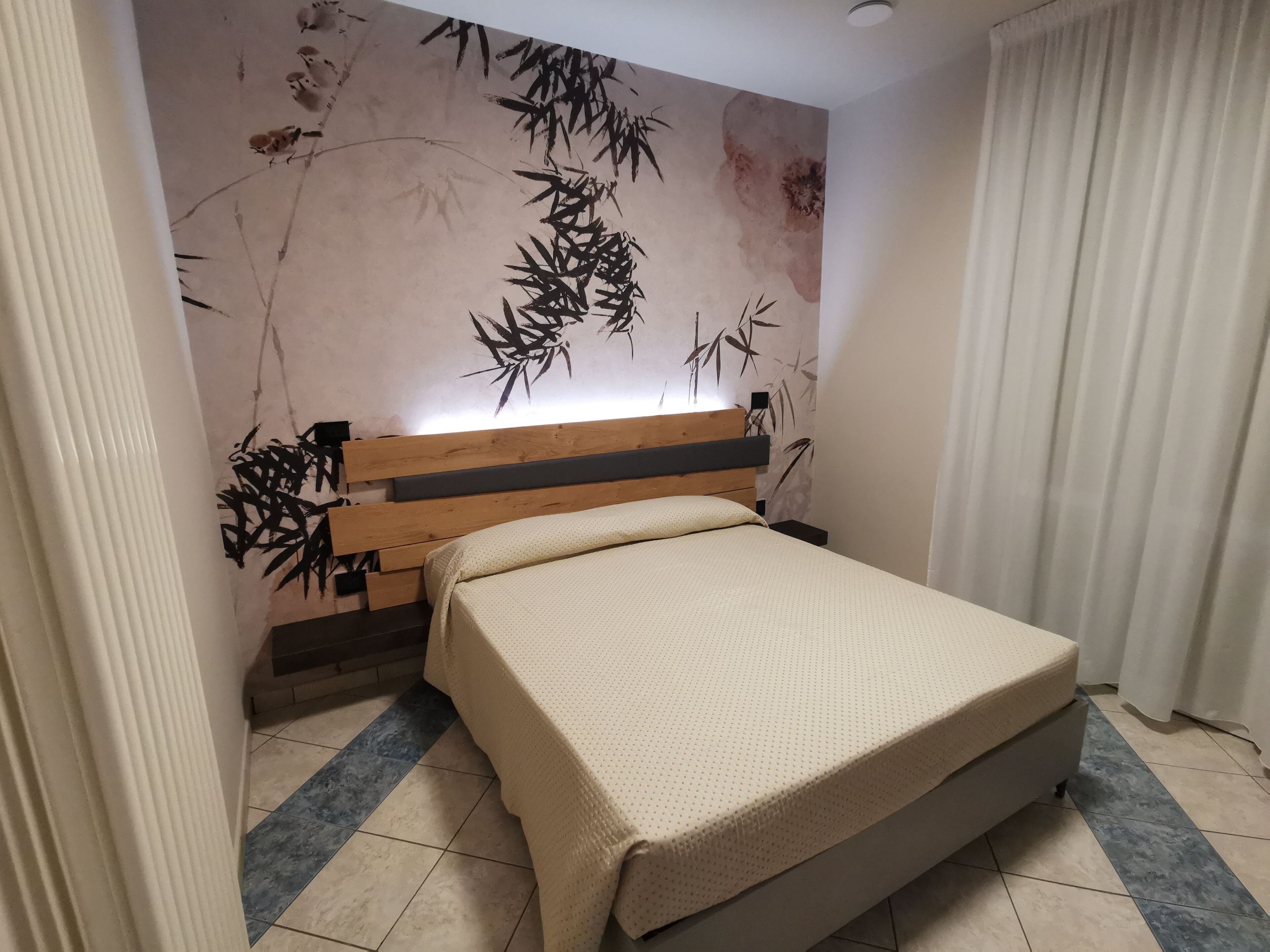 Quarto com cabeceira de madeira, cama com colcha bege e parede com impressão de bambu.