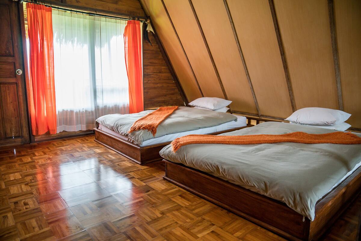 Quarto com duas camas de madeira, piso de madeira e uma janela grande com cortinas.