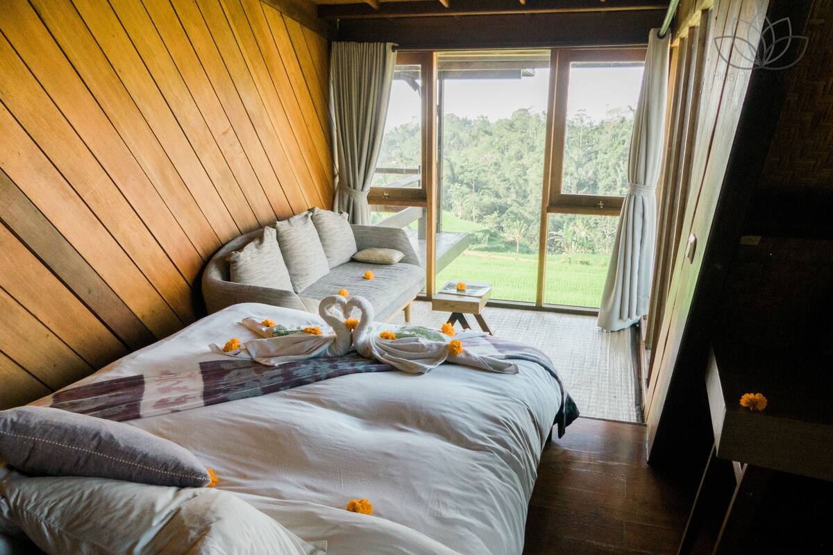 Quarto com cama, sofá e uma janela grande com vista para o verde.