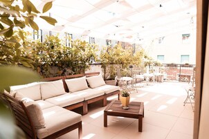 Pátio iluminado pelo sol com um sofá de madeira e mesa de centro, rodeado por plantas em vasos e móveis de jardim.