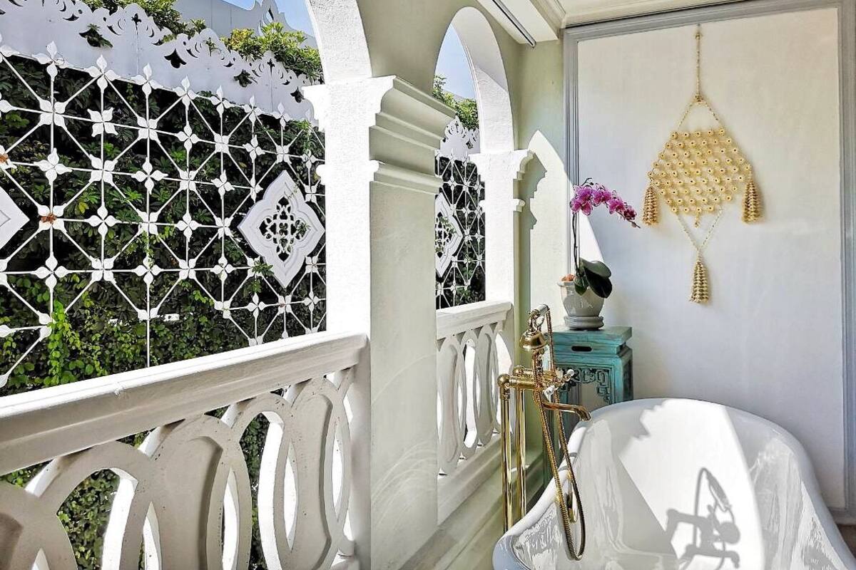 Varanda branca e ornamentada com grade de metal geométrica, uma banheira dourada e um enfeite pendente decorativo.