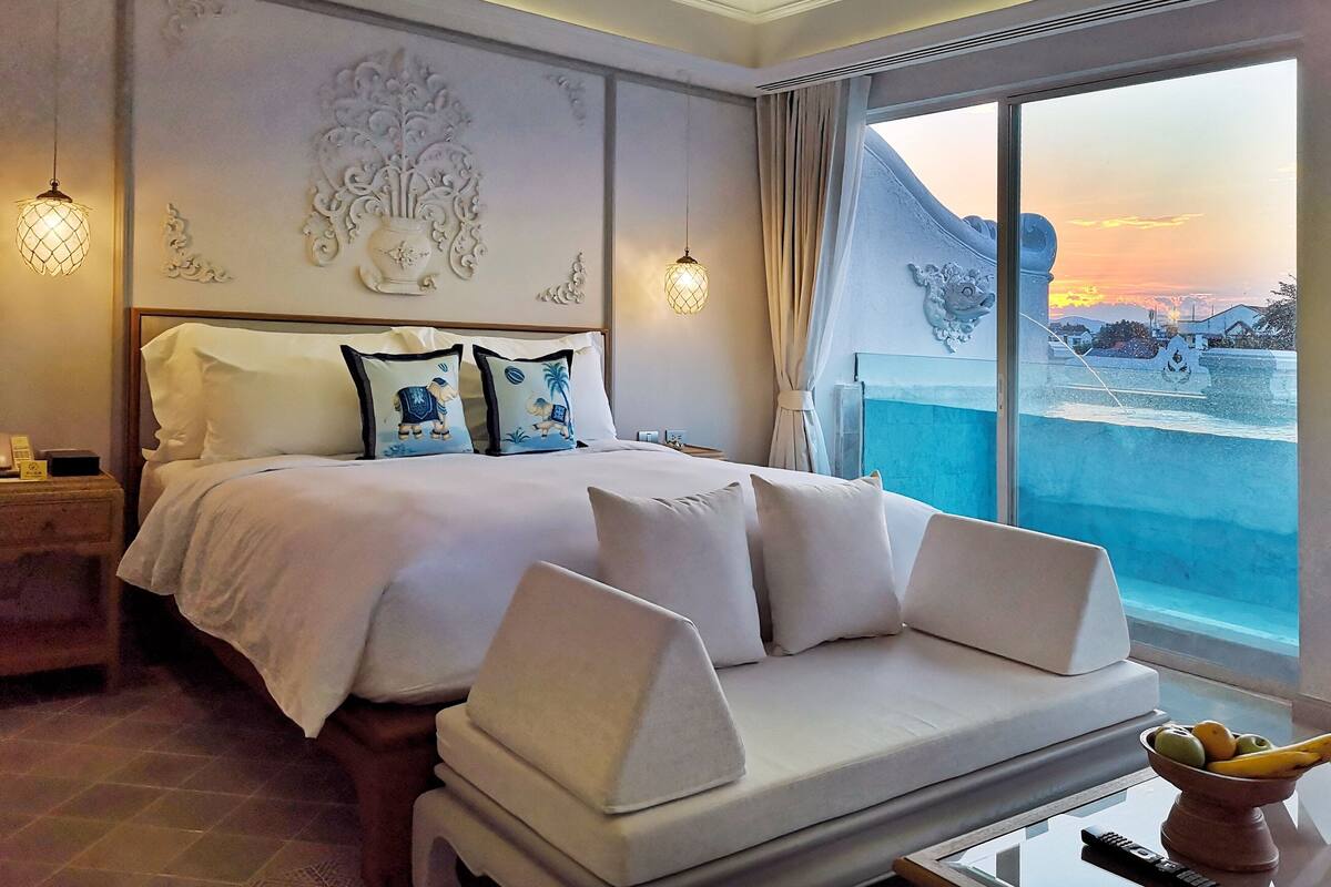 Quarto de hotel com cama grande, sofá, mesa de cabeceira e vista para uma piscina ao pôr do sol.