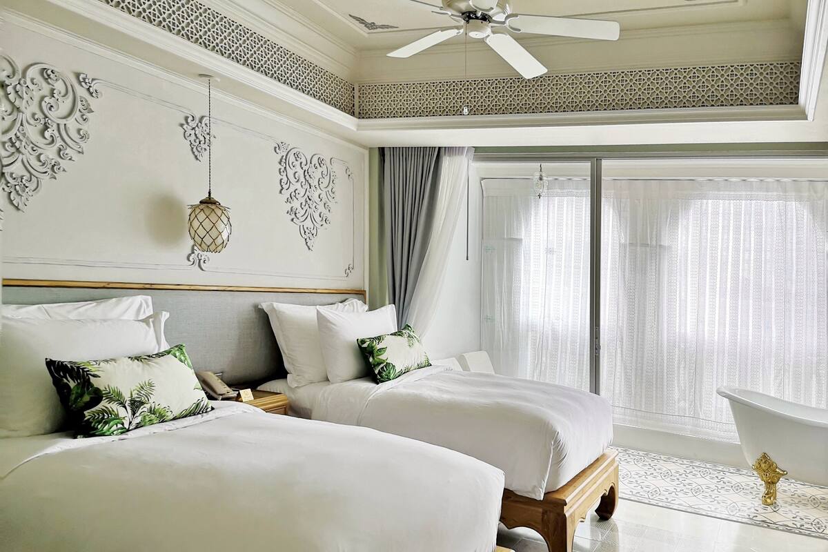 Quarto de hotel com duas camas, ventoinha no teto, banheira e uma janela grande com cortinas translúcidas.