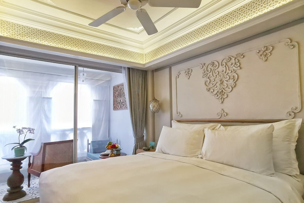 Quarto de hotel com uma cama grande, um ventilador de teto, um painel decorativo na parede, uma janela com cortinas e uma pequena mesa com uma planta.