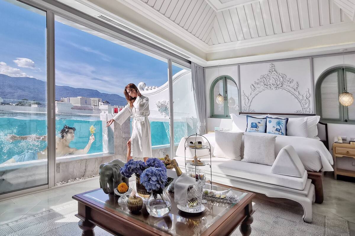 Quarto luxuoso com uma grande janela com vista para uma piscina e montanhas, uma cama com roupa de cama branca e uma mesa de centro com objetos decorativos.