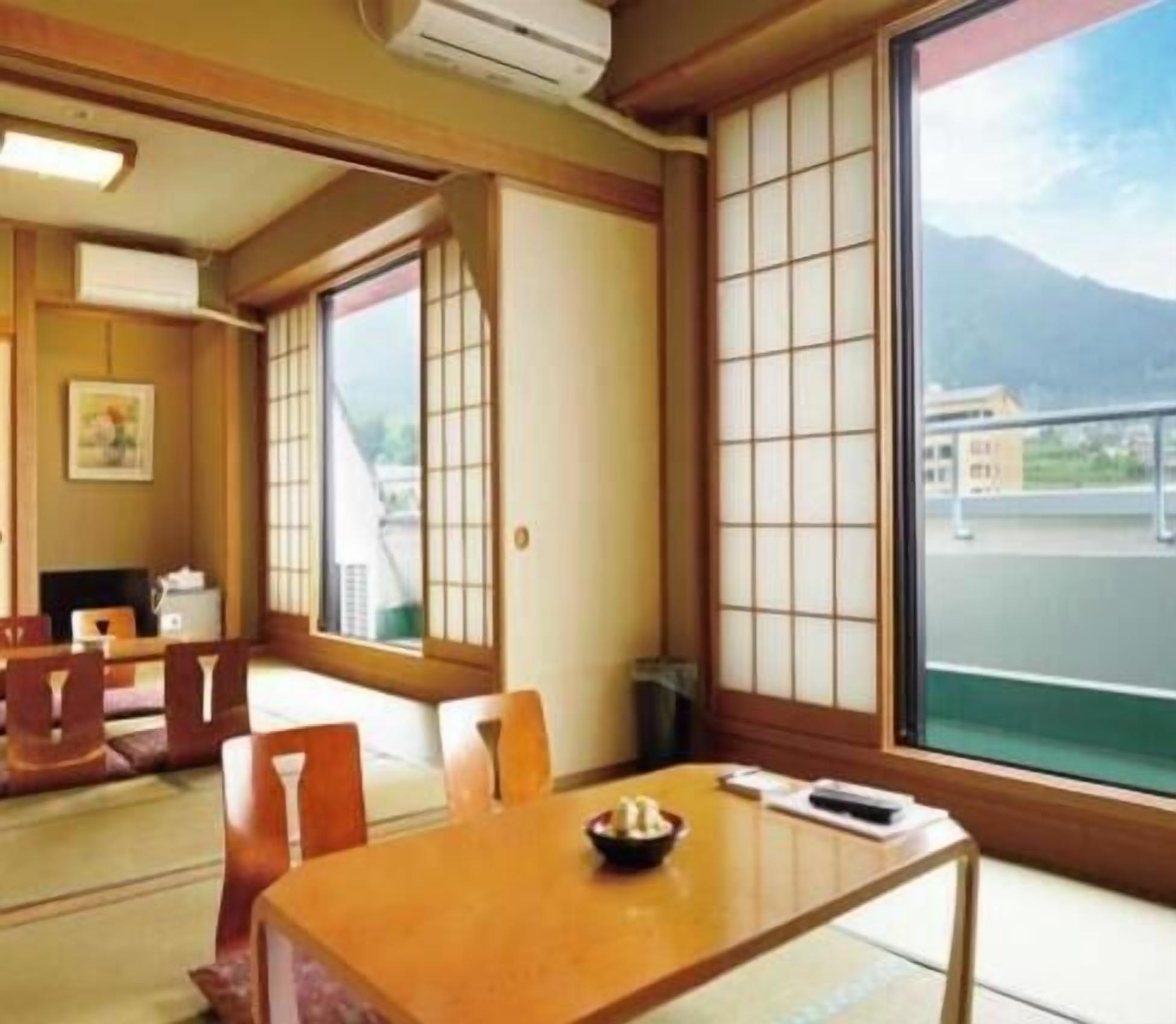 Um quarto japonês tradicional com tatames, uma mesa baixa com uma tigela, e grandes janelas com vista para uma montanha.