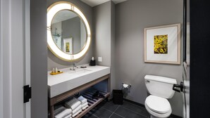 Casa de banho moderna com lavatório branco, espelho redondo com luz, sanita e quadro emoldurado na parede.