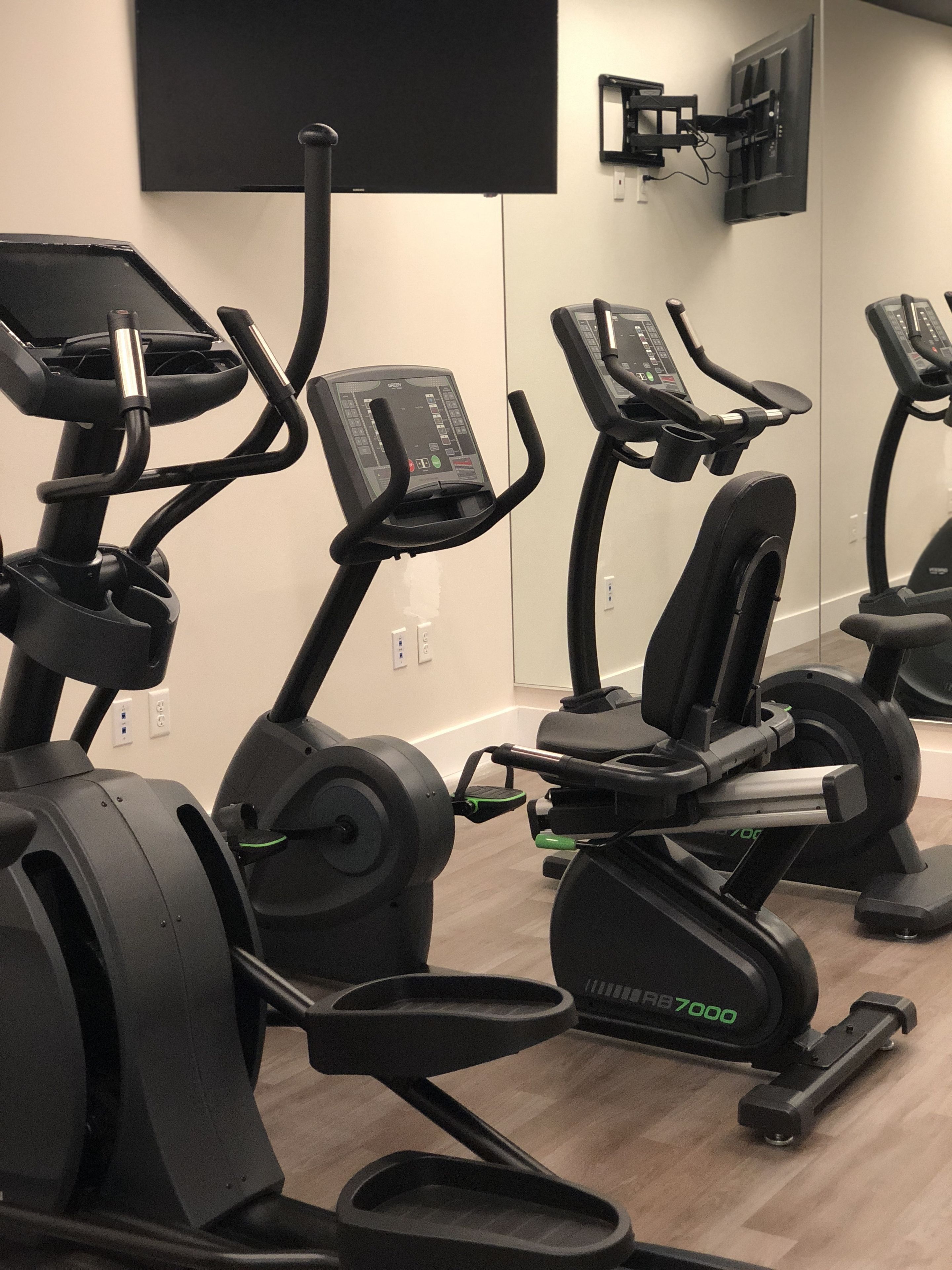 Academia com várias bicicletas ergométricas, uma televisão fixada na parede e piso de madeira.