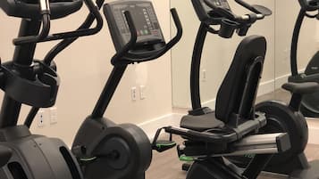 Academia com várias bicicletas ergométricas, uma televisão fixada na parede e piso de madeira.