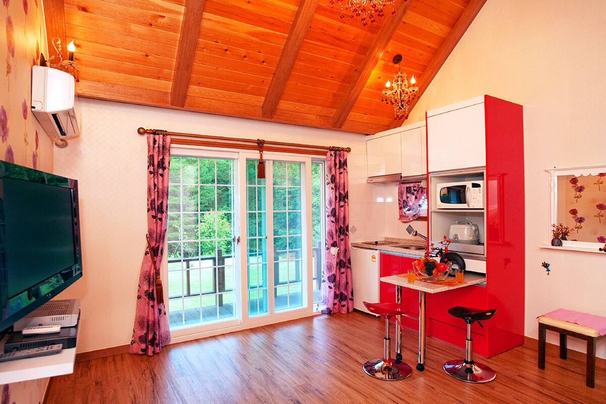 Quarto com teto de madeira, televisão, cozinha equipada com micro-ondas e mesa de jantar com duas cadeiras.