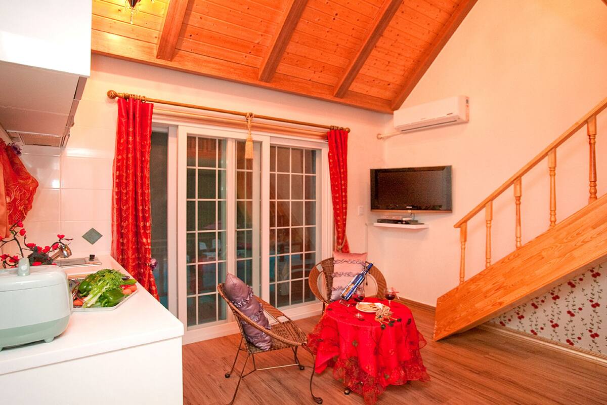 Um quarto com escada de madeira, televisão na parede, mesa de jantar com toalha vermelha e janela com cortinas vermelhas.