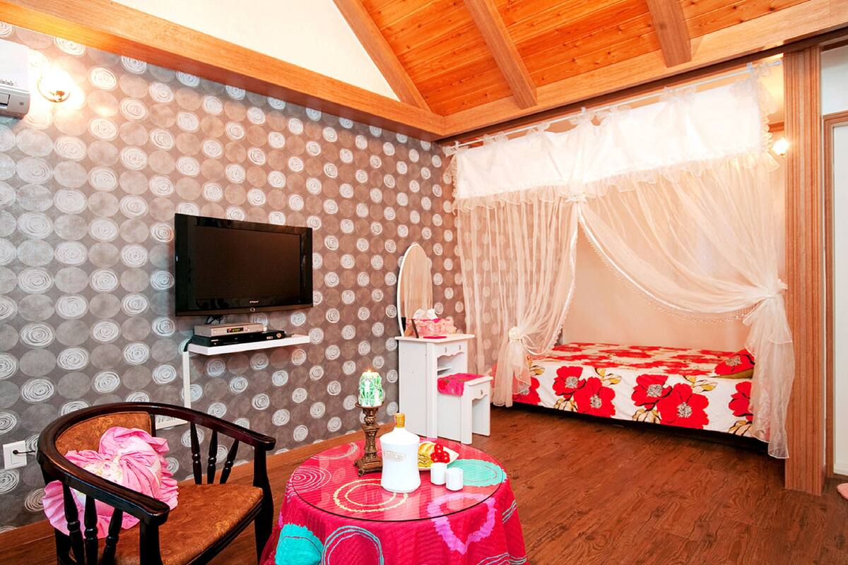 Quarto com televisão, uma cadeira, mesa com tampo de vidro, cama com colcha florida e um espelho.
