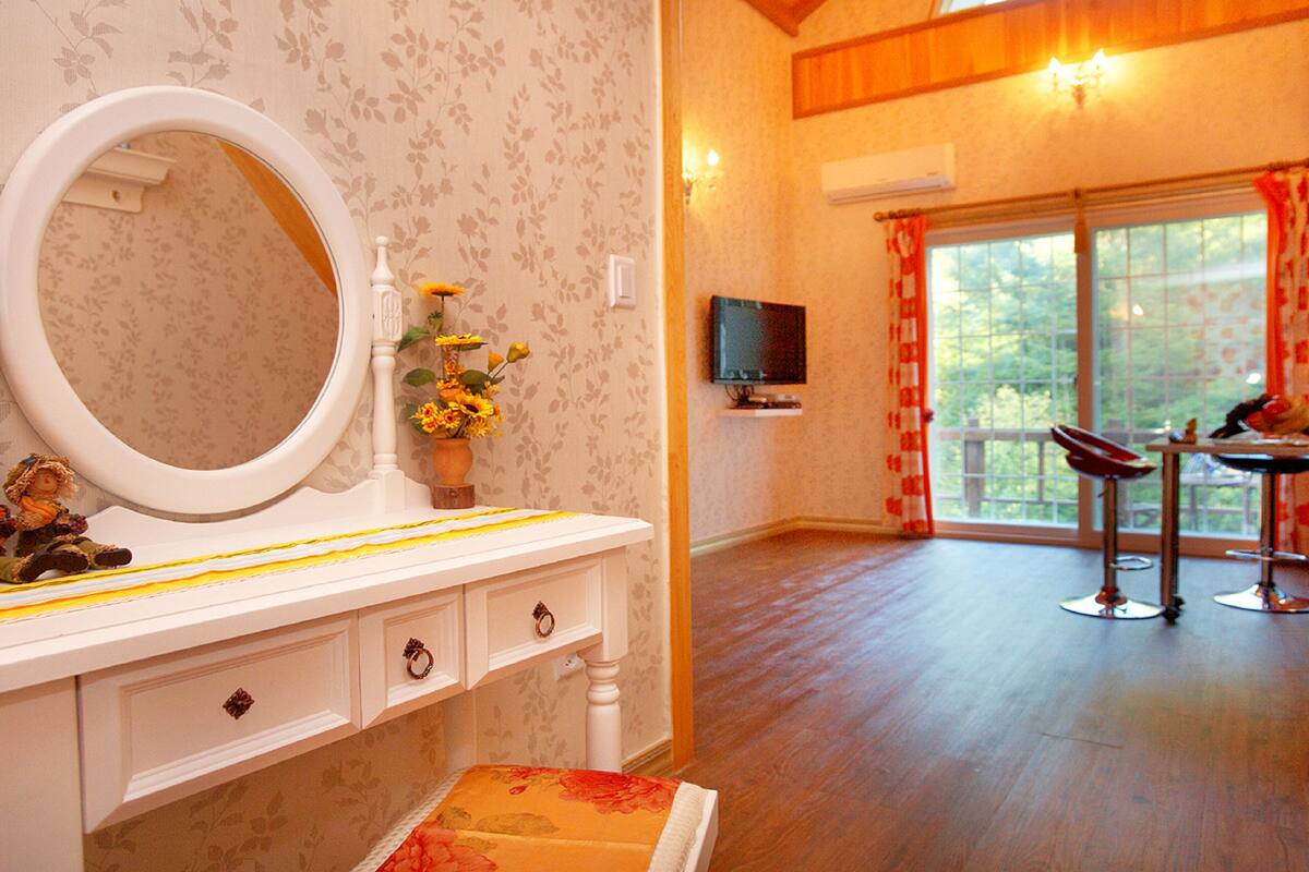 Quarto com penteadeira, televisão e área de refeições com vista para o exterior.