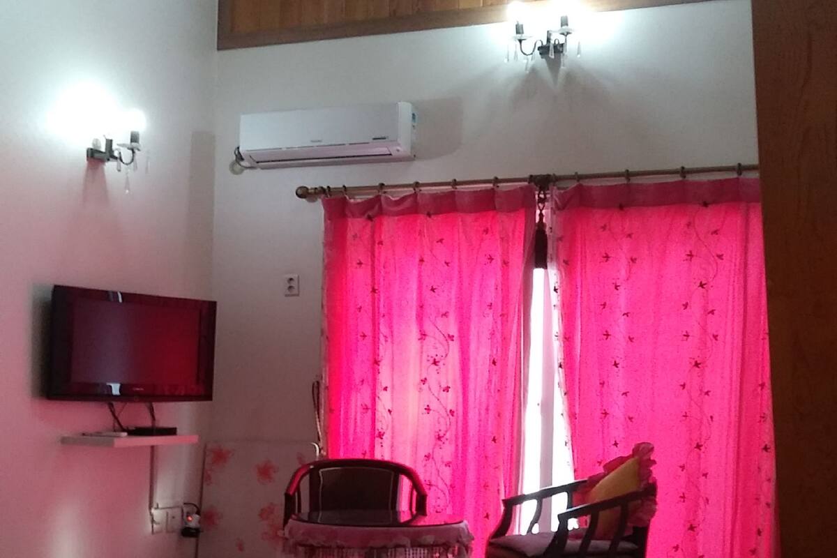 Uma sala de estar com teto de madeira, uma televisão sobre um suporte, duas cadeiras, uma mesinha e uma janela com cortinas cor-de-rosa.