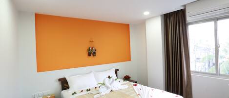 Quarto de hotel com uma parede de destaque laranja, uma cama bem arrumada com lençóis brancos e pétalas de rosa, uma pequena mesa com telefone e uma janela com cortinas.