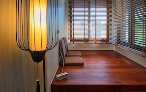 Um quarto com piso de madeira, um banco, uma luminária e uma janela com persianas.