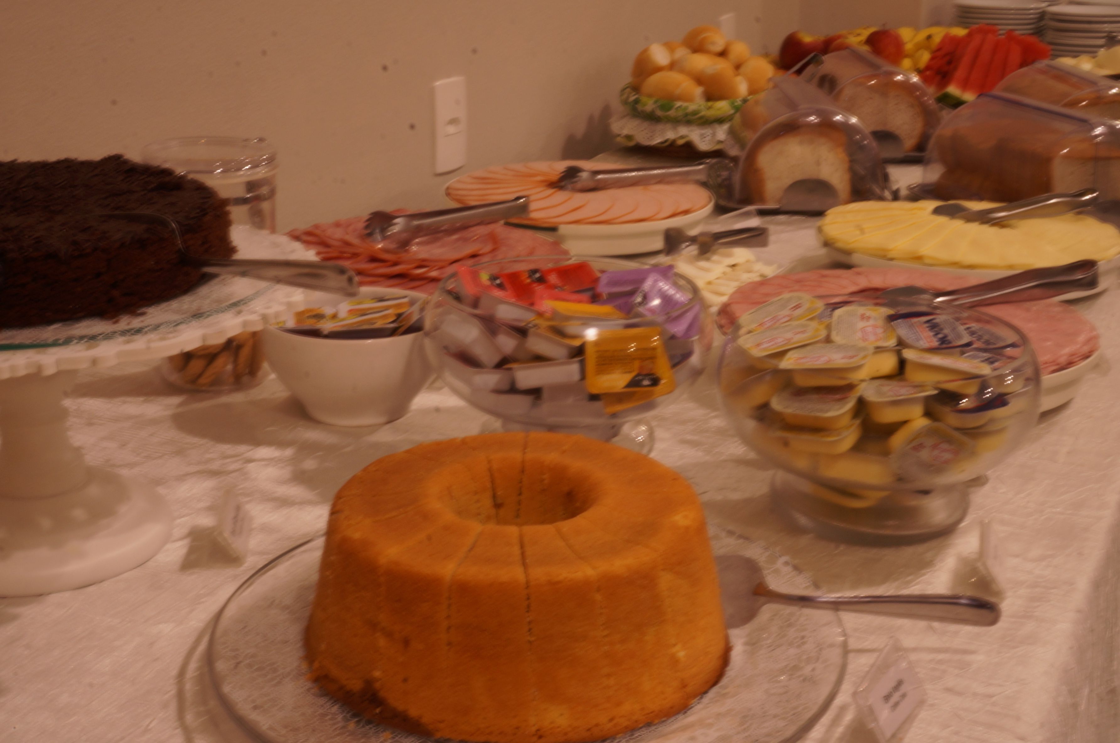 Mesa de buffet com diversos alimentos, incluindo um bolo bundt, queijos, frutas e outras sobremesas.