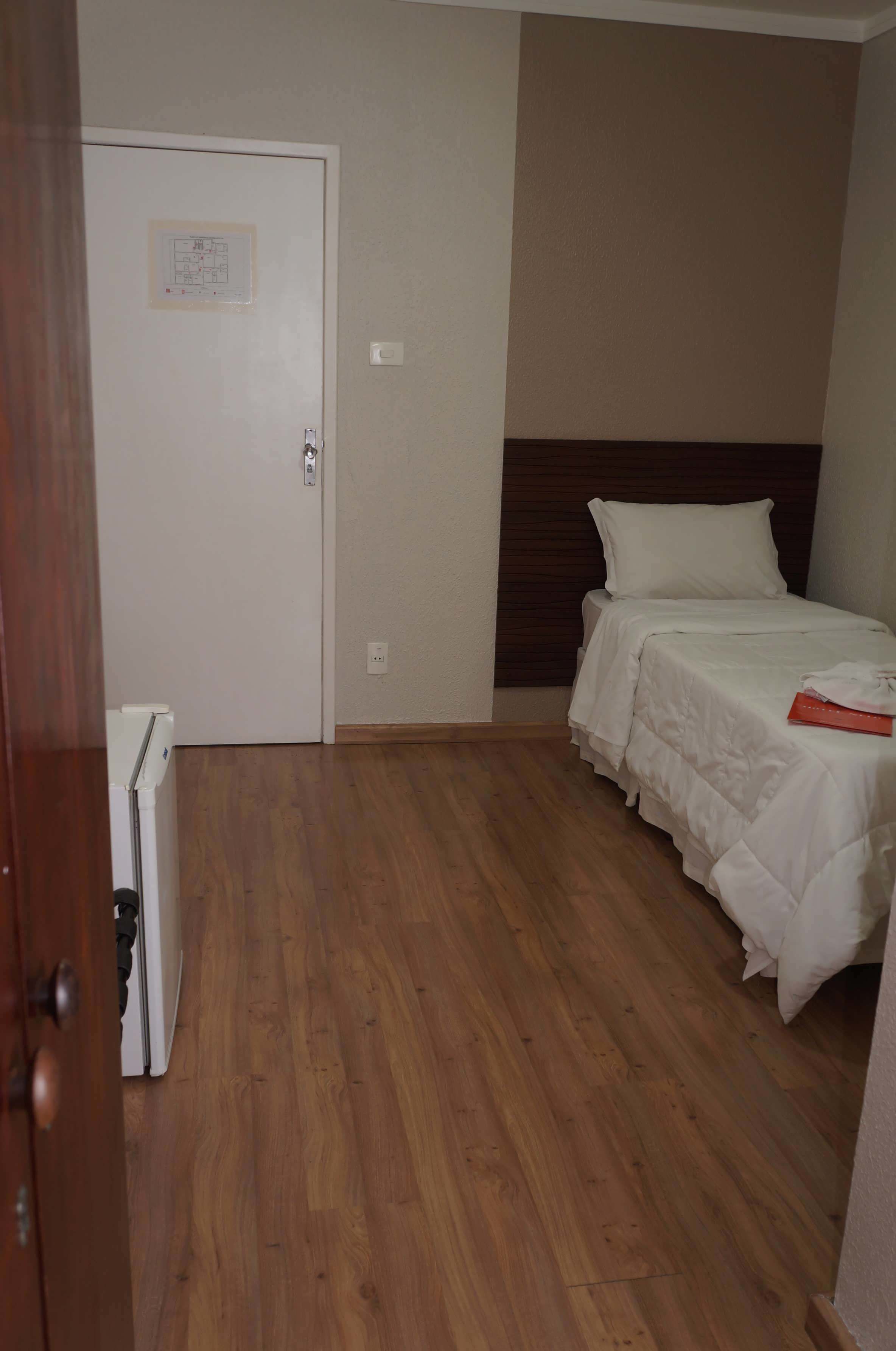 Um quarto com piso de madeira, uma cama branca com cabeceira e uma porta com um aviso.